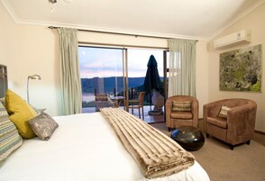 Quarto de hotel com uma cama grande, duas poltronas, uma mesa e vista para o exterior.