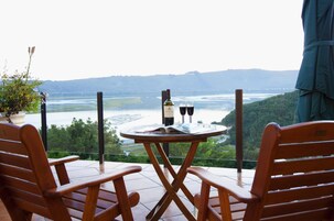 Uma mesa de madeira com uma garrafa de vinho e duas taças numa varanda com vista para um rio e colinas.