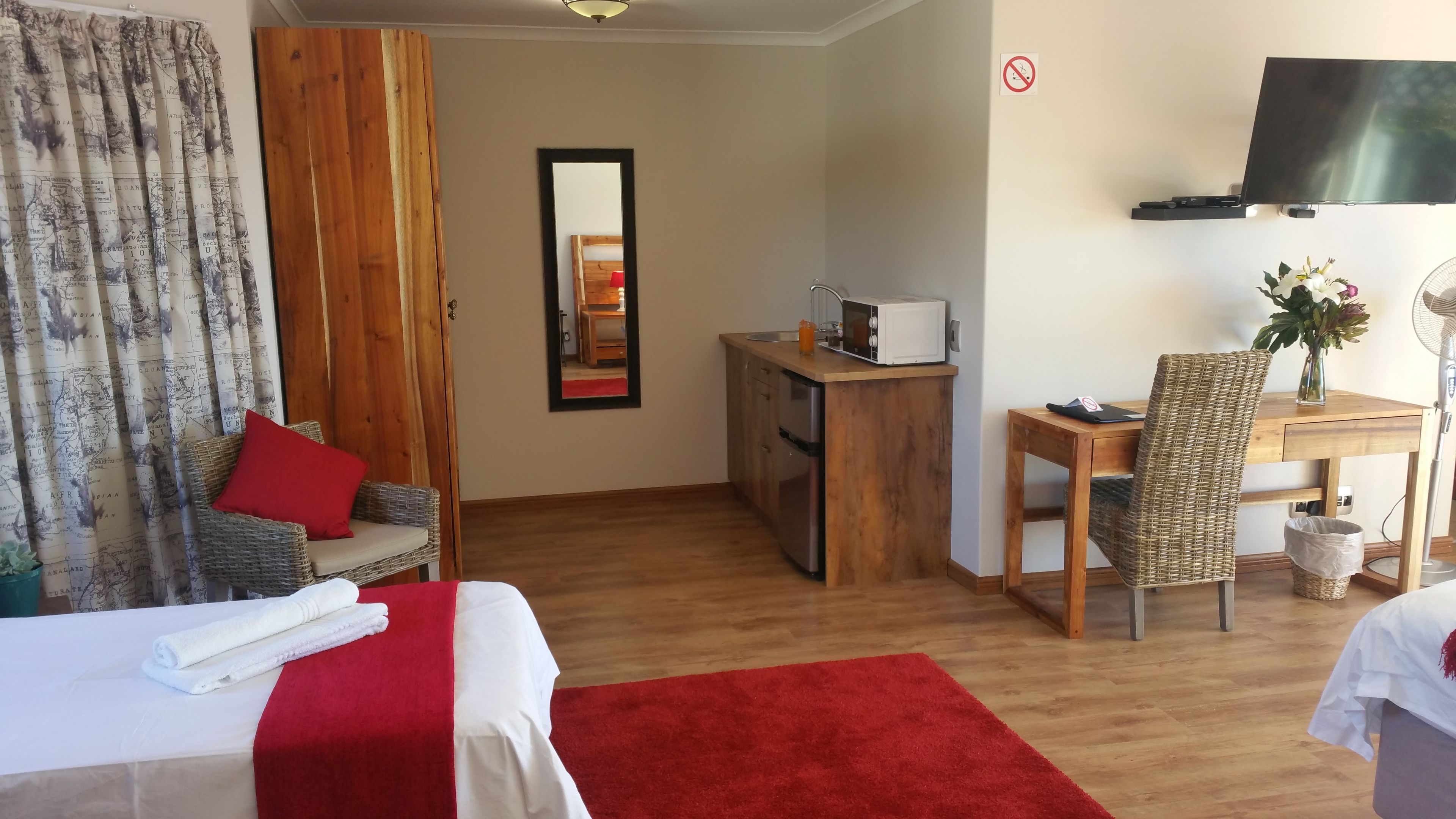 Quarto de hotel com cama, tapete vermelho, escrivaninha de madeira, cadeira, televisão e espelho.