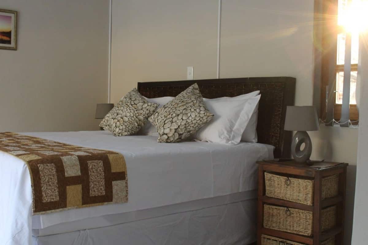 Cama bem arrumada com colcha estampada, duas almofadas decorativas, luminária de cabeceira e mesa de cabeceira de madeira pequena com cestos.