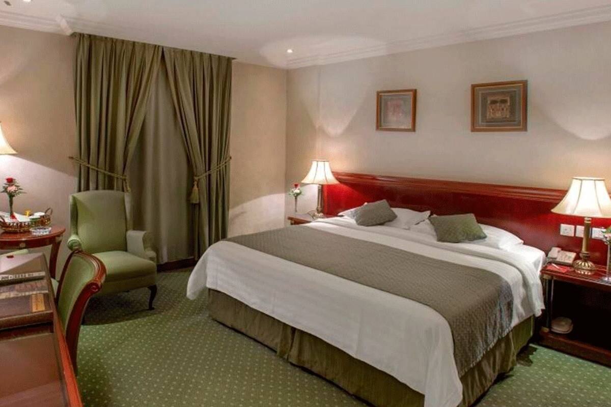 Quarto de hotel com uma cama grande, duas mesas de cabeceira com luminárias, uma cadeira, uma escrivaninha com luminária e uma televisão.