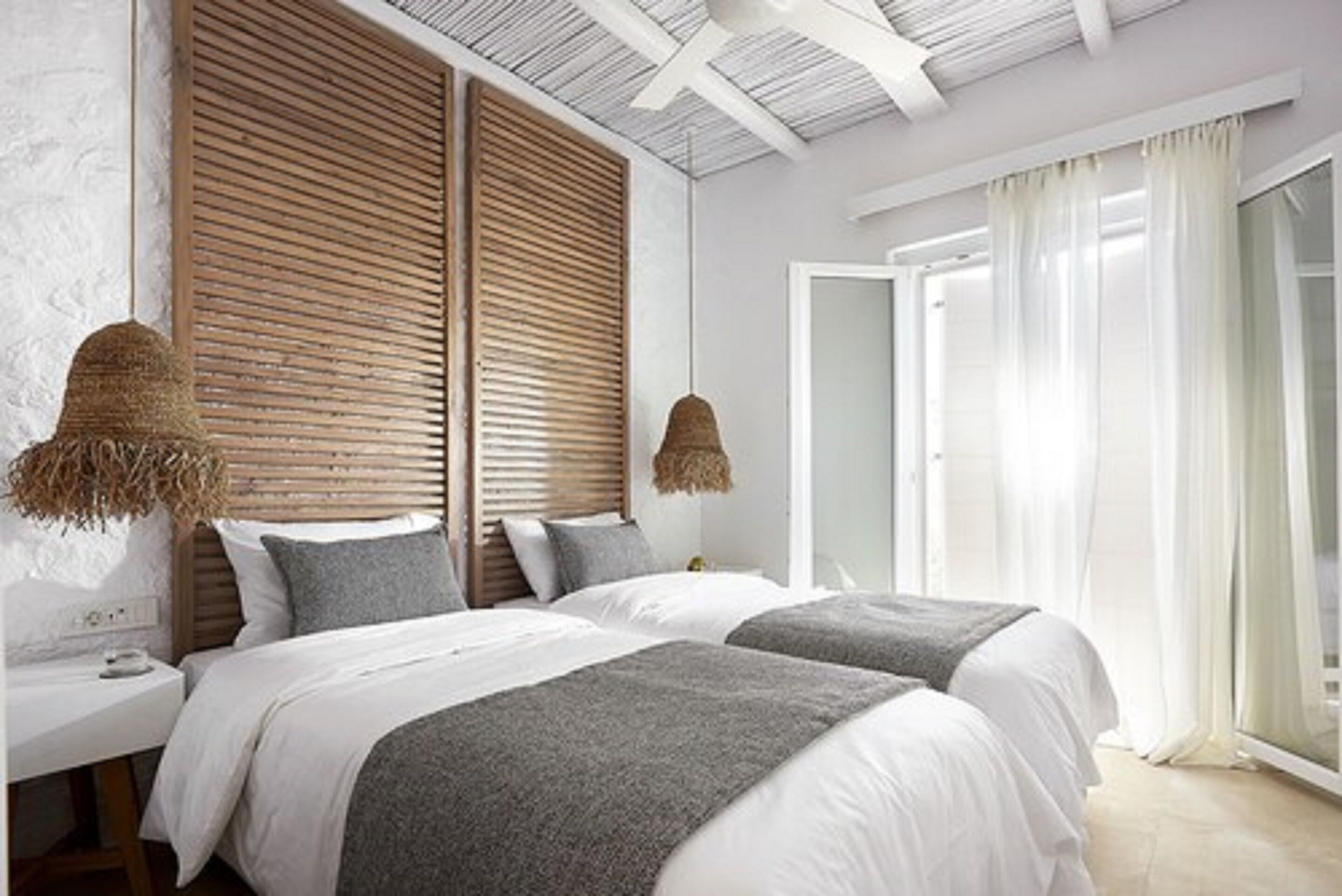 Quarto com duas camas, cabeceira de madeira, mesa de cabeceira branca e uma janela grande com cortinas leves.
