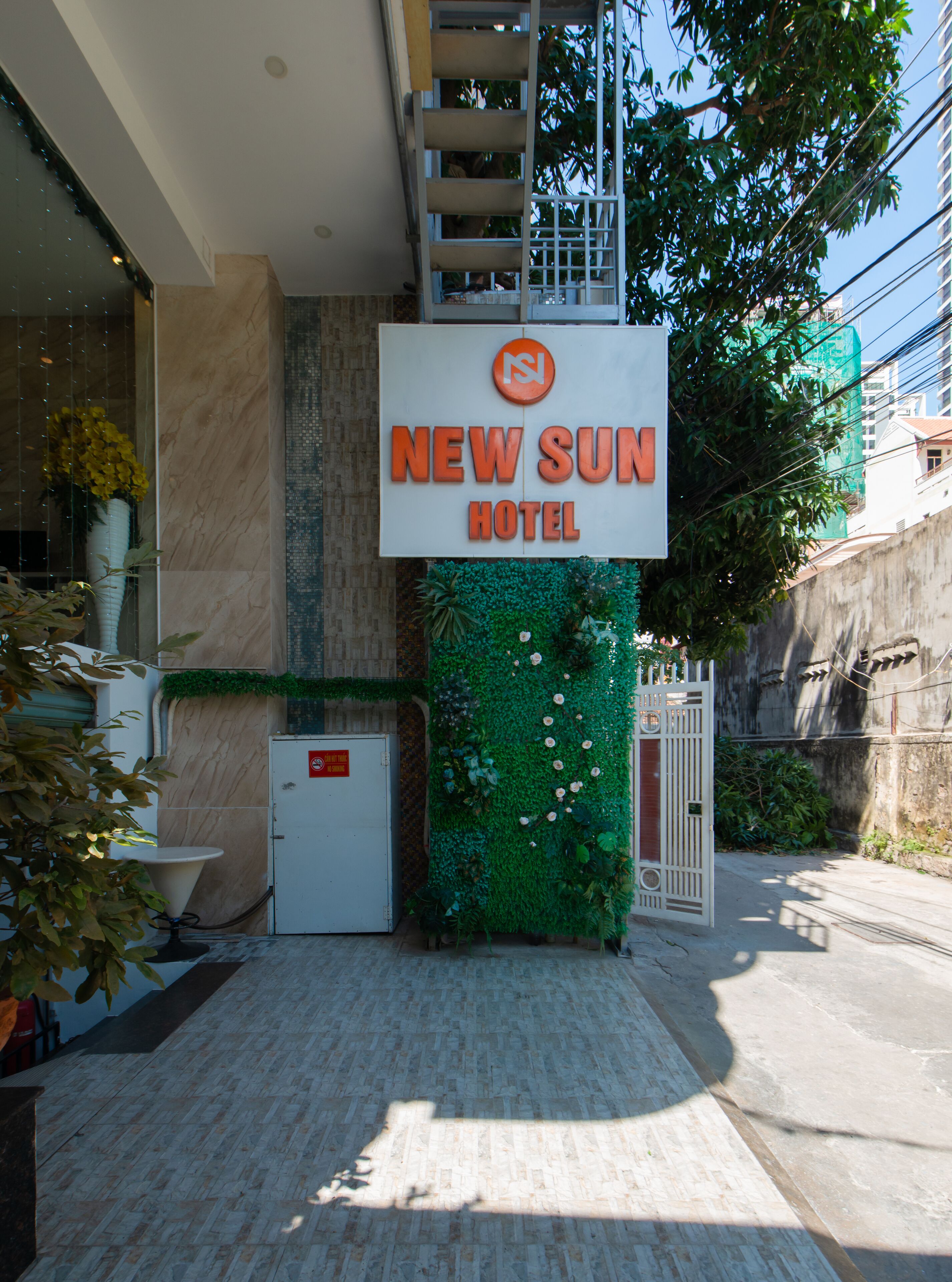 Exterior do Hotel New Sun, com uma placa, um pequeno portão de entrada e uma escadaria que leva à entrada.