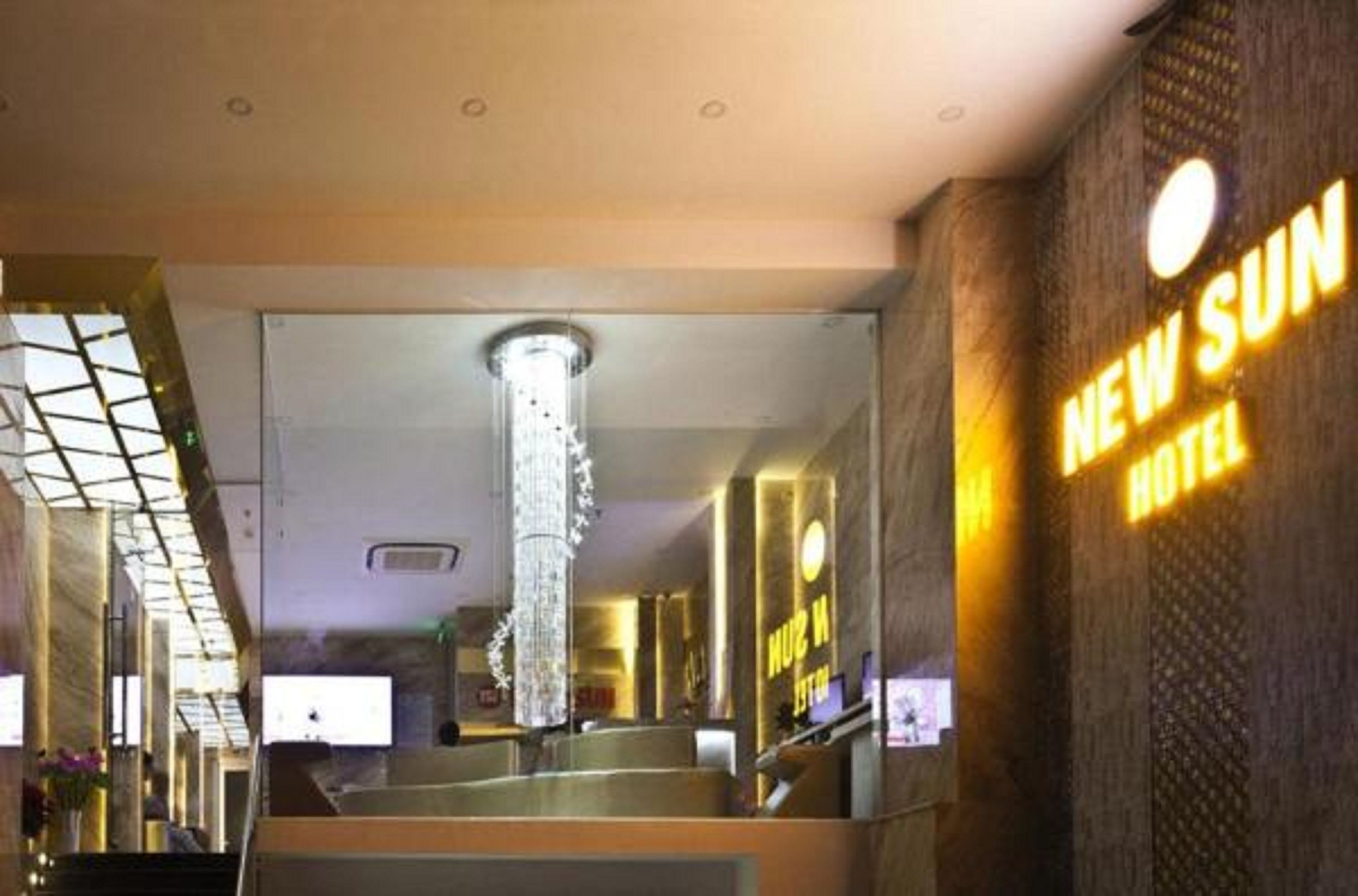 Um hall de hotel com uma escadaria moderna, um lustre grande e uma placa com a inscrição 'NEW SUN HOTEL'.