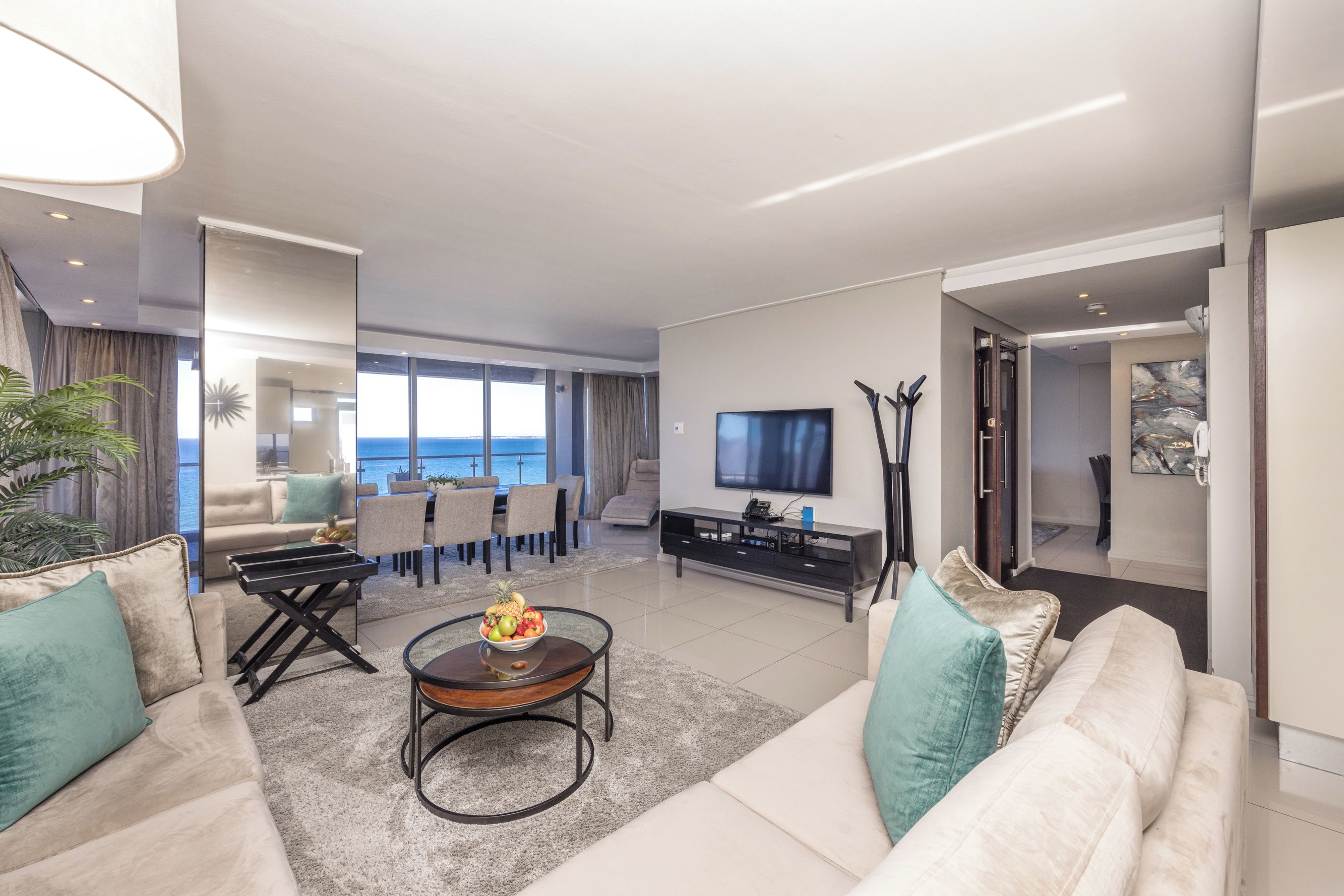 Uma sala de estar moderna com um sofá, uma mesa de centro com frutas, uma televisão e vista para o mar através de grandes janelas.