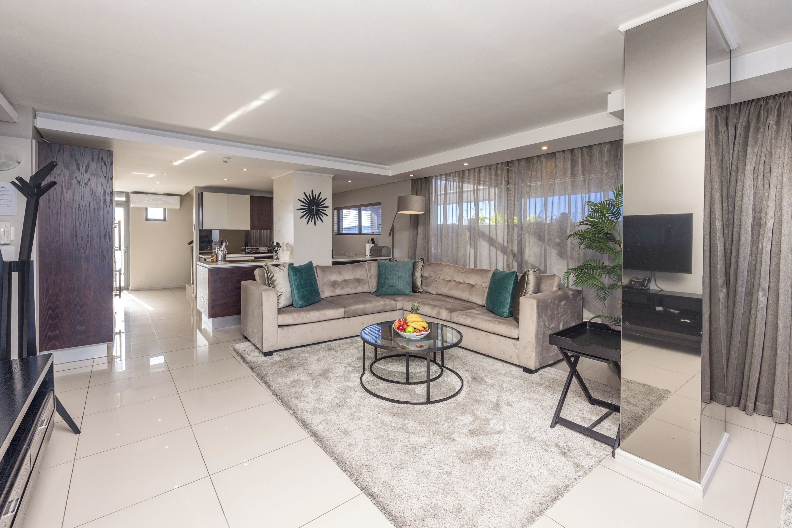Uma sala de estar moderna com um sofá seccional, uma mesa de centro com frutas, uma TV e uma cozinha ao fundo.