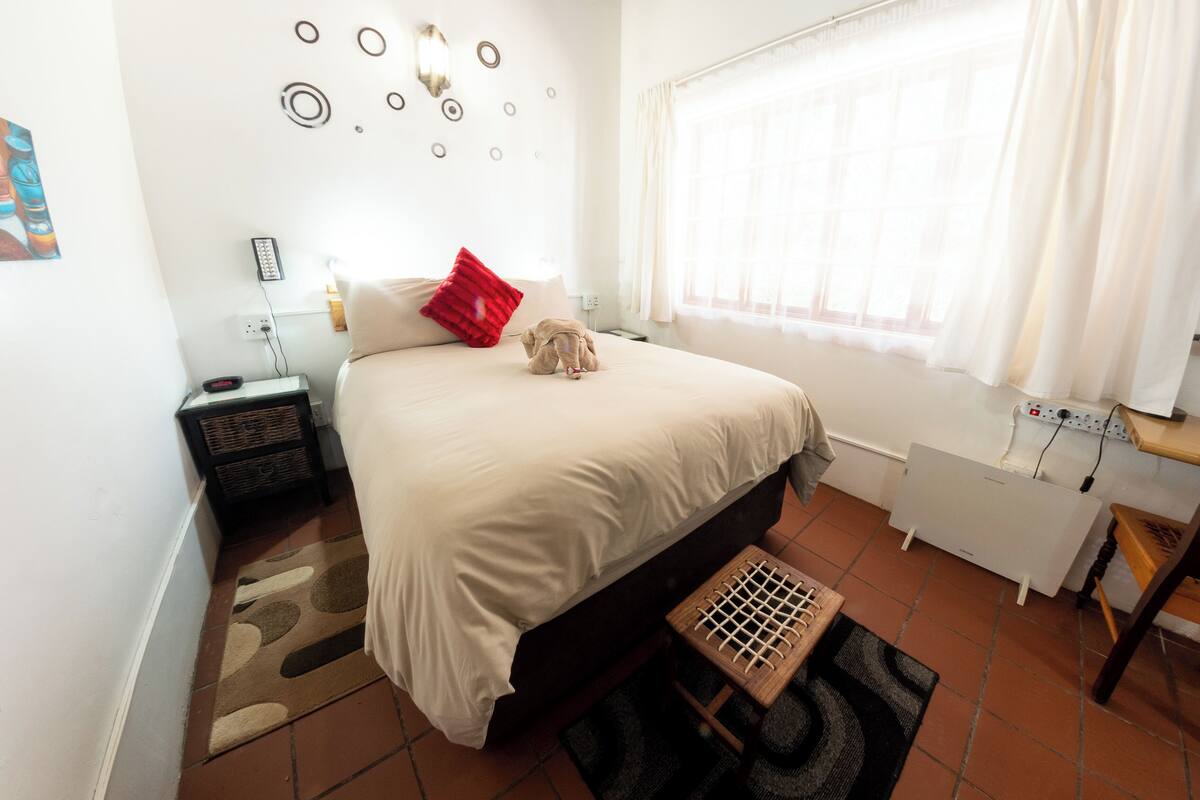 Quarto com cama, mesa de cabeceira, mesinha, janela com cortinas e parede decorada com elementos circulares.