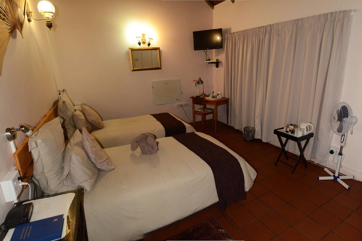 Quarto de hotel com duas camas, uma escrivaninha, televisão, ventoinha e janela com cortinas.
