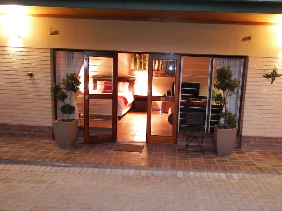 Entrada de hotel com porta de vidro que dá para um quarto com cama e televisão. Há plantas em vasos do lado de fora da porta.