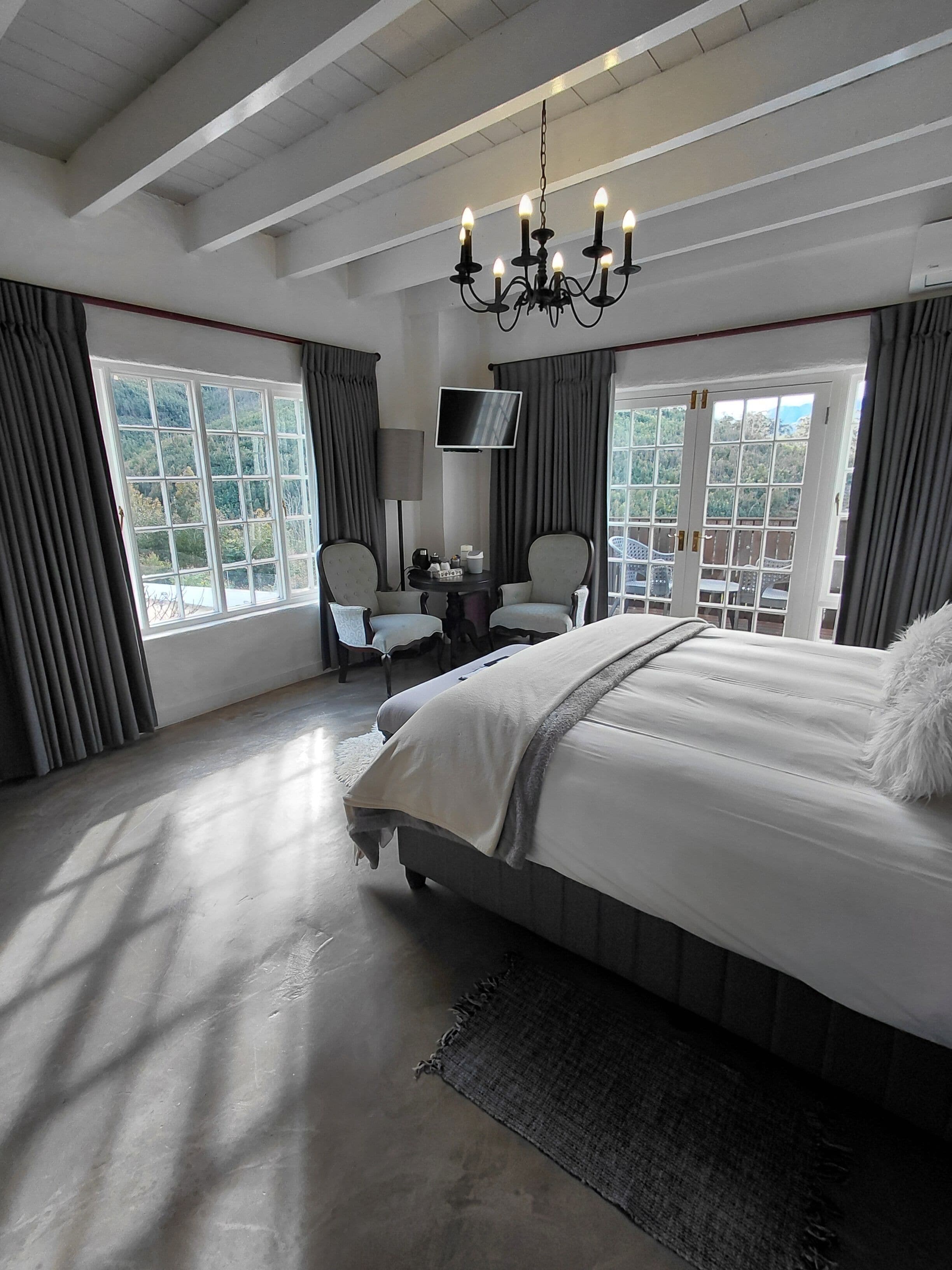 Quarto de hotel com uma cama grande, duas cadeiras, uma escrivaninha, uma televisão e janelas amplas.