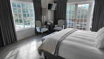 Quarto de hotel com uma cama grande, duas cadeiras, uma escrivaninha, uma televisão e janelas amplas.