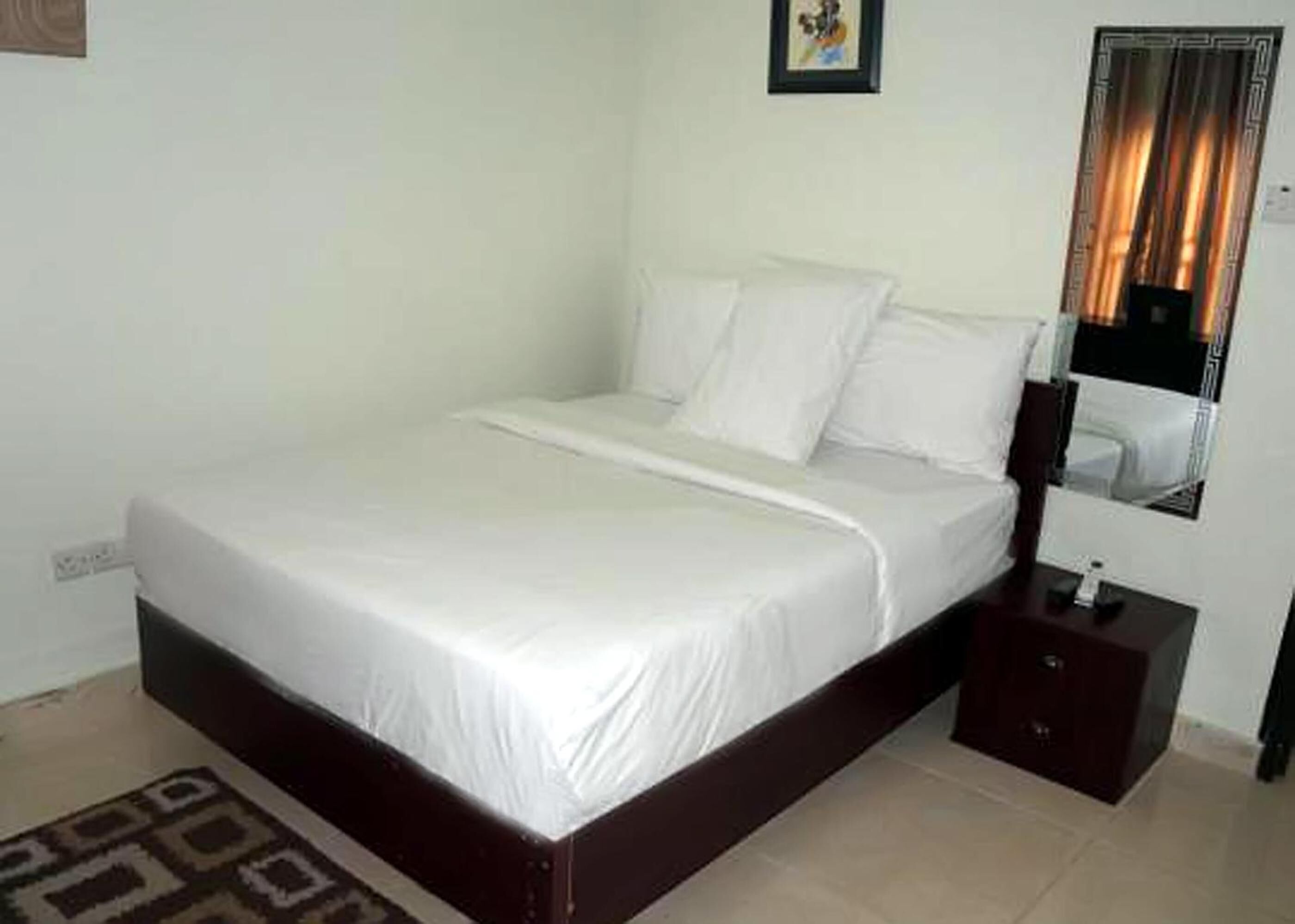 Cama bem arrumada com lençóis brancos em quarto simples, com cabeceira de madeira escura e uma pequena mesa de cabeceira.