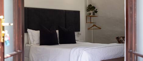 Uma cama bem arrumada com roupa de cama branca e duas almofadas pretas, uma mesinha de cabeceira com uma planta e um banco de madeira.