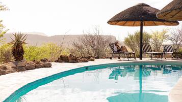 Uma área de piscina com espreguiçadeiras e guarda-sóis, rodeada por vegetação de deserto e pedras.