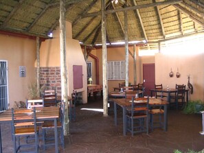 Uma área de refeições rústica com mesas e cadeiras de madeira, parede de tijolos e telhado de palha.