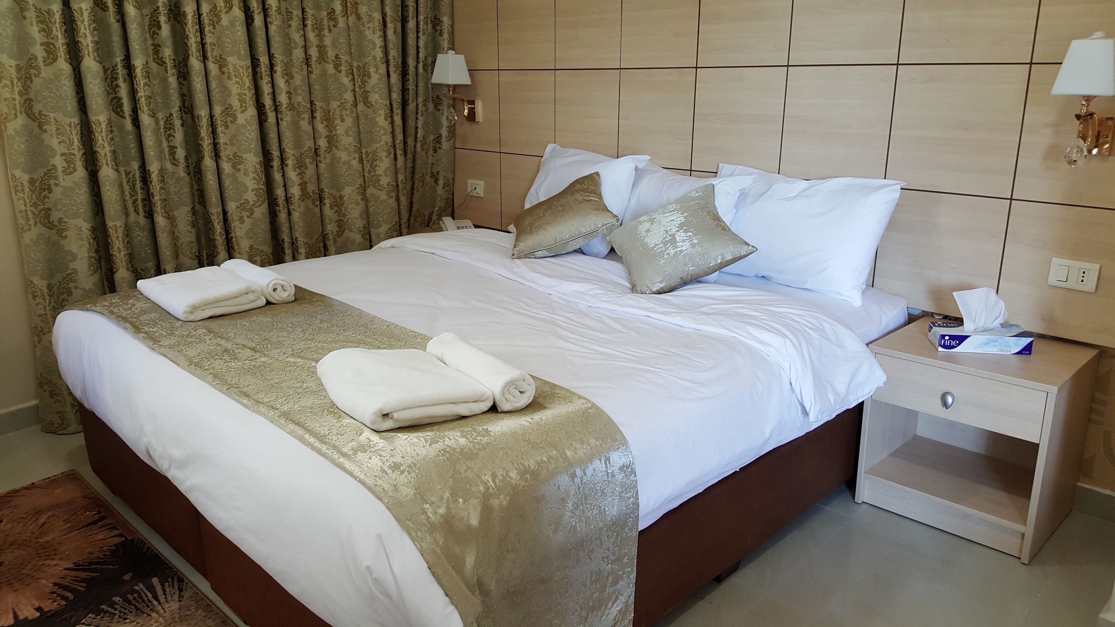 Cama bem arrumada com lençóis brancos, um xale dourado e duas toalhas enroladas ao lado. Há uma mesa de cabeceira com um porta-lenços e uma luminária. O quarto tem uma cabeceira de madeira e uma cortina com padrão floral.