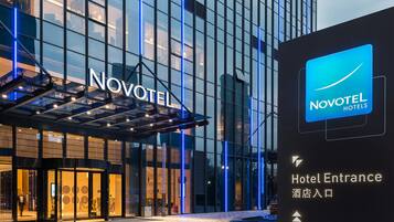 Entrada de um hotel Novotel com fachada de vidro, sinalização iluminada e placa indicando estacionamento.