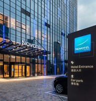 Entrada de um hotel Novotel com fachada de vidro, sinalização iluminada e placa indicando estacionamento.