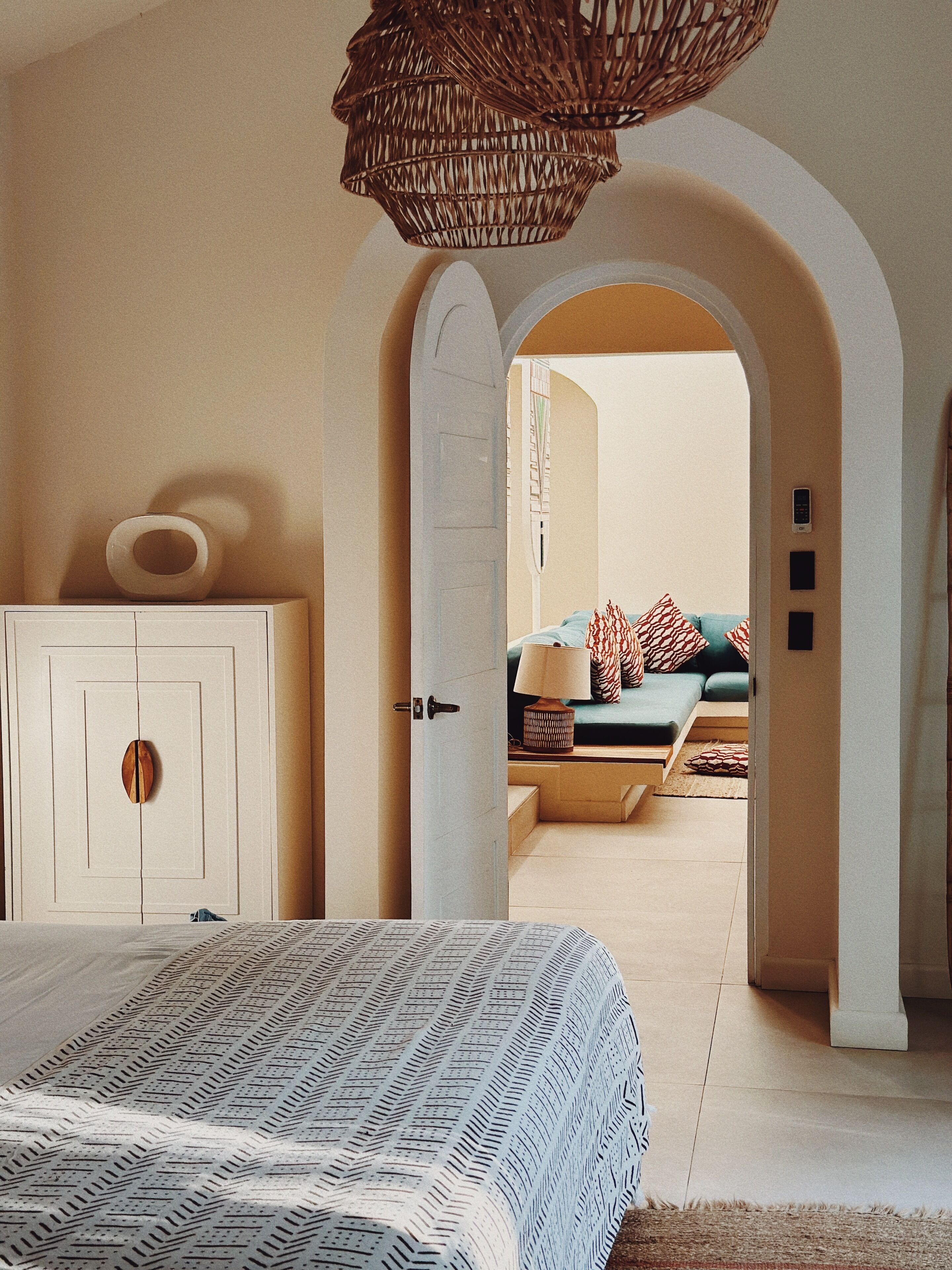 Quarto com cama, guarda-roupa e porta que dá para outro ambiente com sofá e abajur.