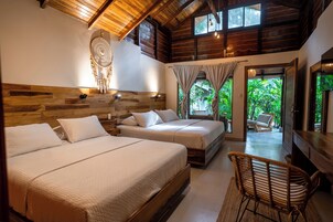 Quarto com duas camas, cabeceira de tecido, um sonho apanhador fixado na parede e vista para o verde pela janela.