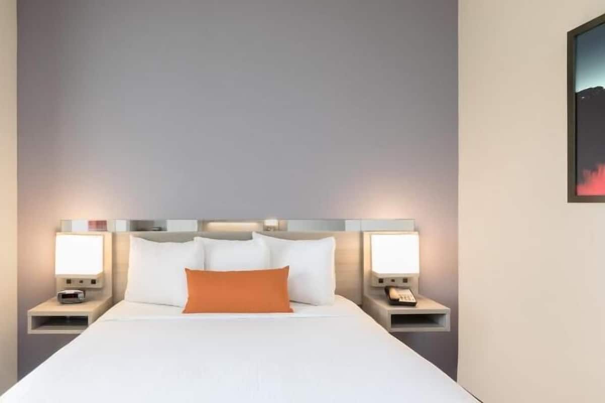 Quarto de hotel com uma cama grande, duas mesinhas de cabeceira, um criado-mudo com abajur e luminária na parede.