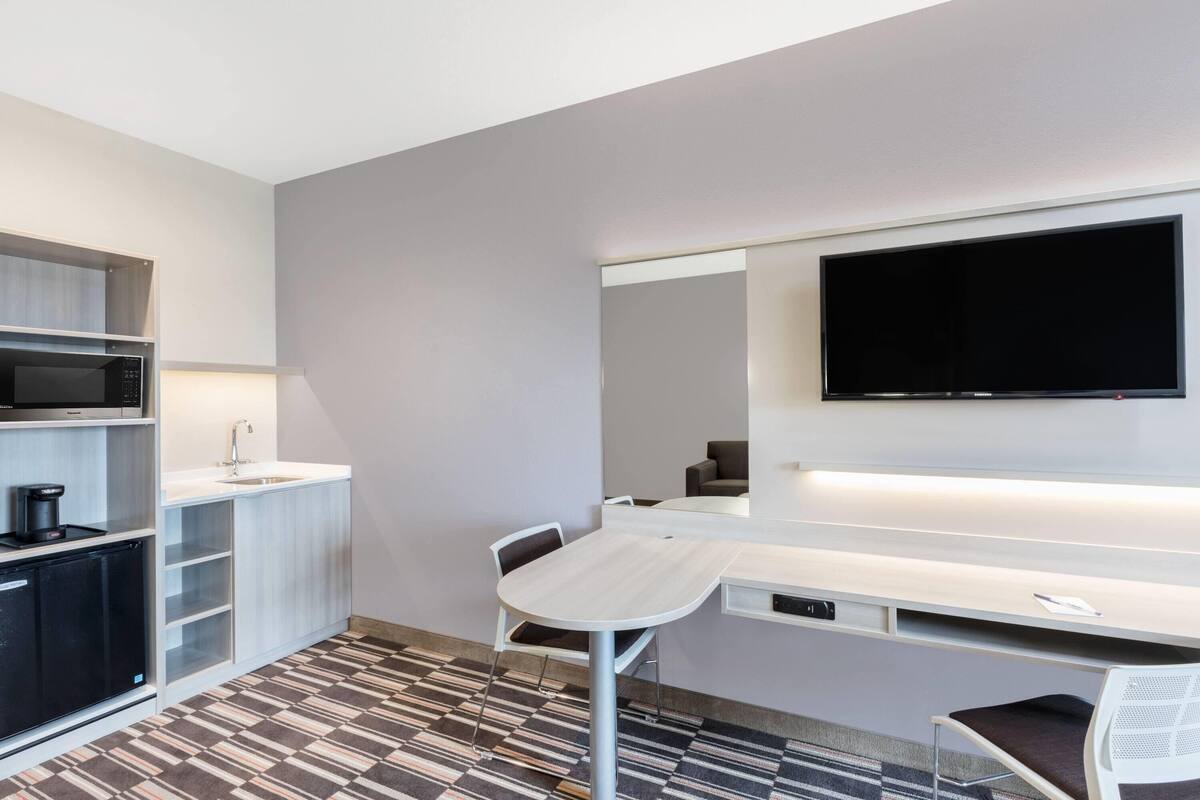 Quarto de hotel moderno com cozinha compacta, televisão de tela plana na parede, mesa de jantar com cadeiras e piso estampado.