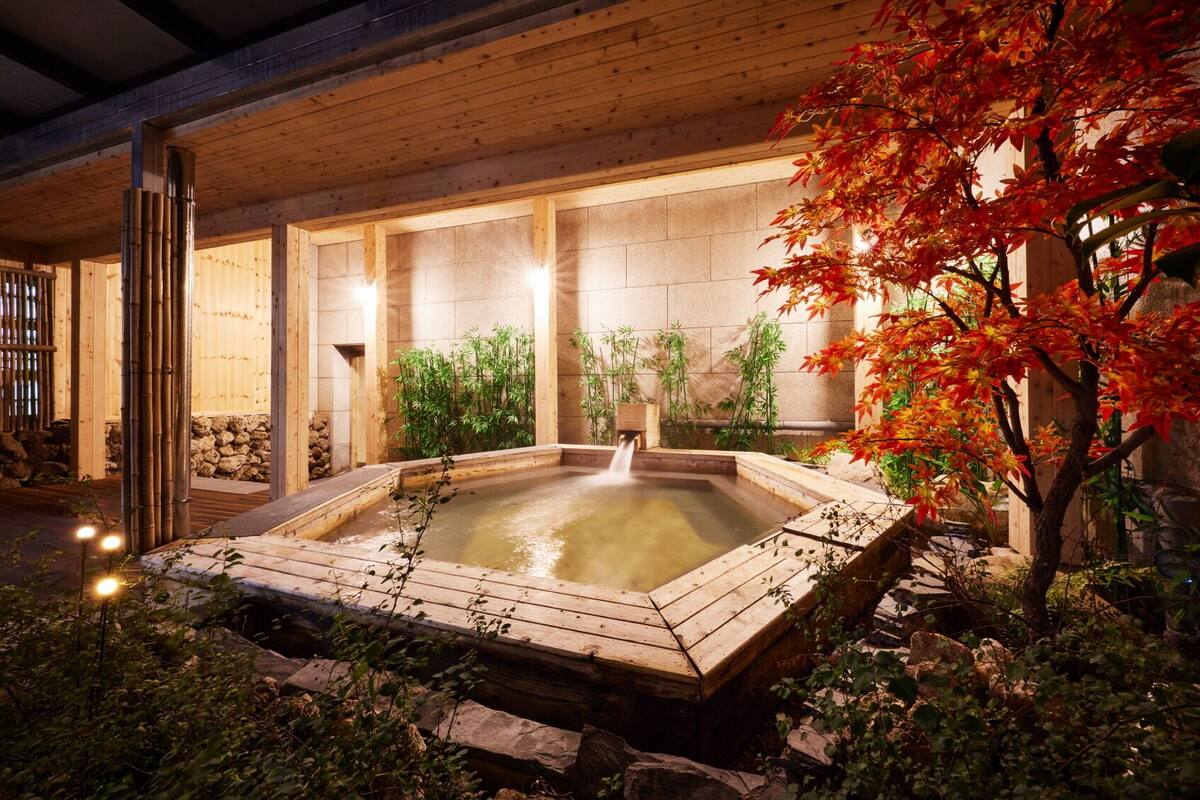 Uma área de águas termais com um deck de madeira, plantas de bambu e um muro de pedra.