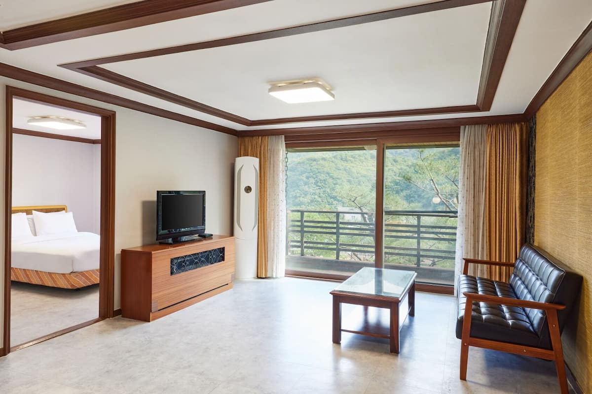 Quarto de hotel com cama, televisão sobre um móvel de madeira, sofá, mesa e varanda com vista para o verde.
