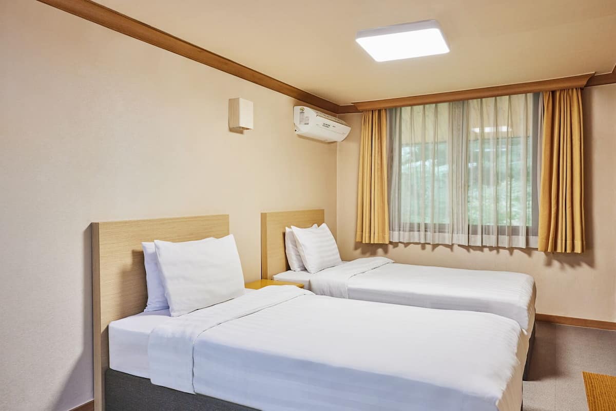 Quarto de hotel com duas camas de solteiro, janela com cortinas finas, ar condicionado e luminária no teto.