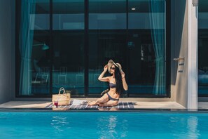 Uma mulher sentada à beira da piscina, com uma bebida e uma bolsa, em frente a um edifício moderno com grandes janelas.