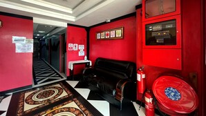 Um lobby de hotel com um sofá de couro preto, paredes vermelhas, um extintor de incêndio e um painel de controle vermelho.