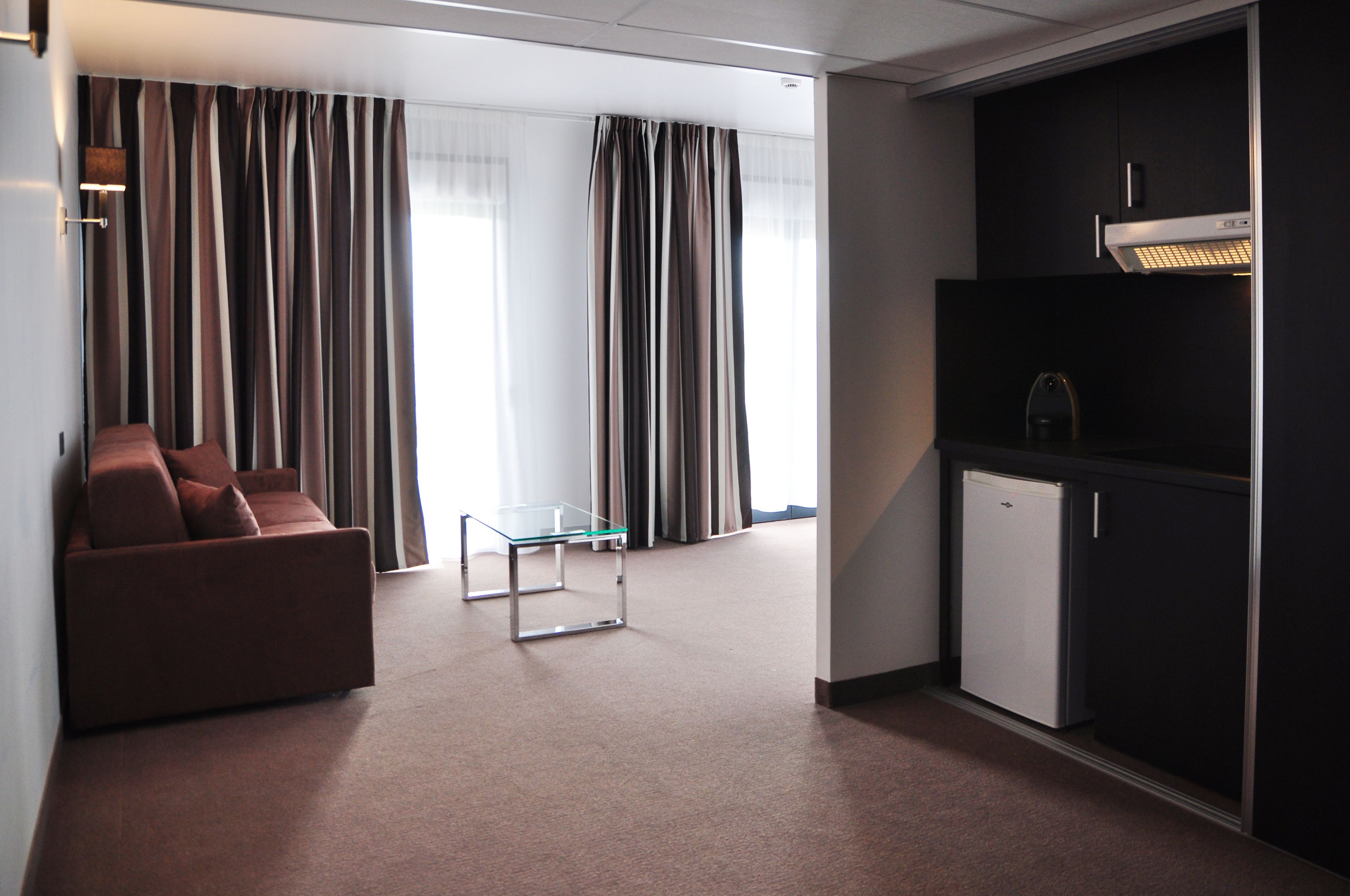 Quarto de hotel moderno com sofá marrom, mesa de centro de vidro, cozinha compacta com frigorífico e cortinas grandes.