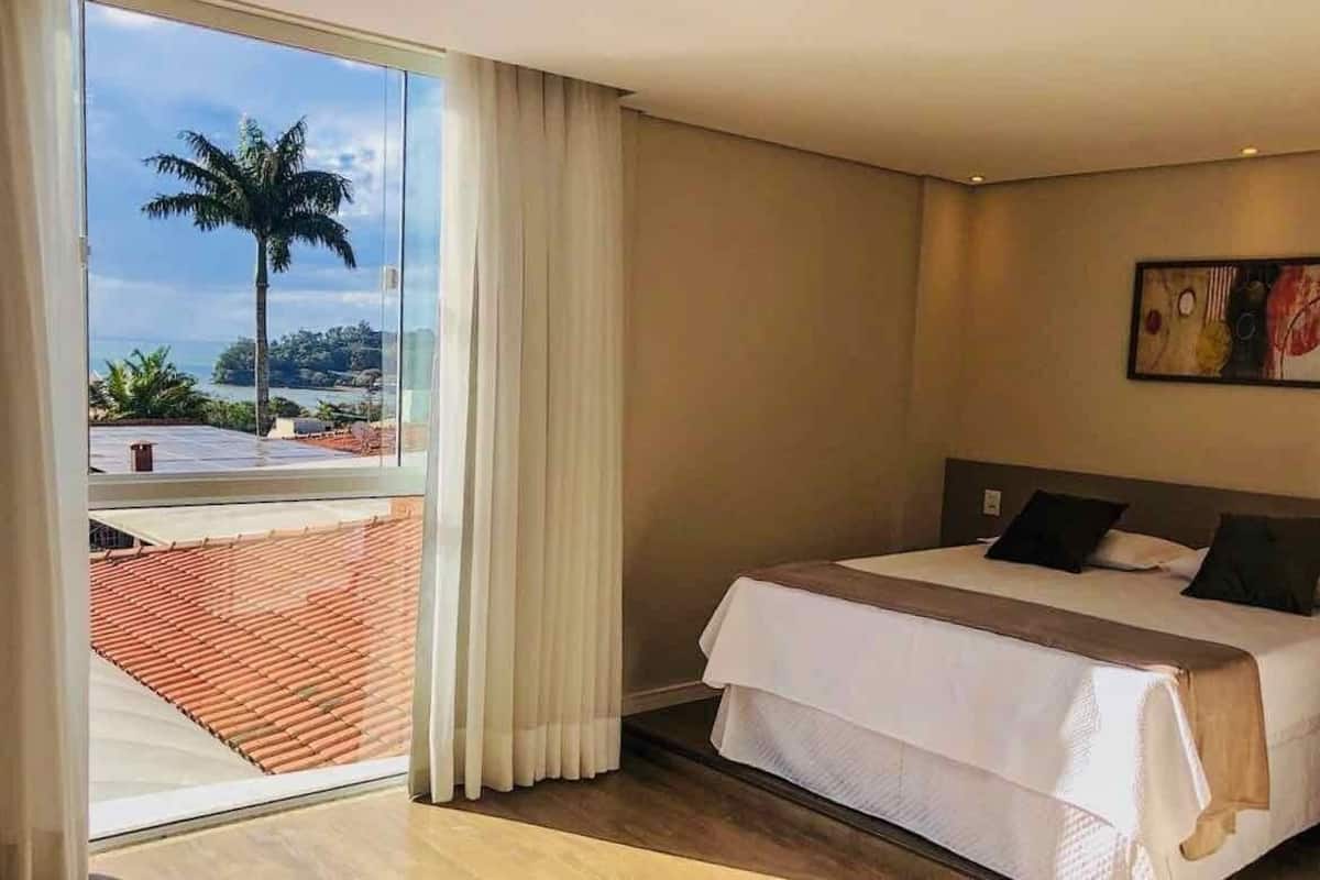 Quarto de hotel com uma cama, um quadro na parede e vista para uma palmeira e um prédio através da janela.