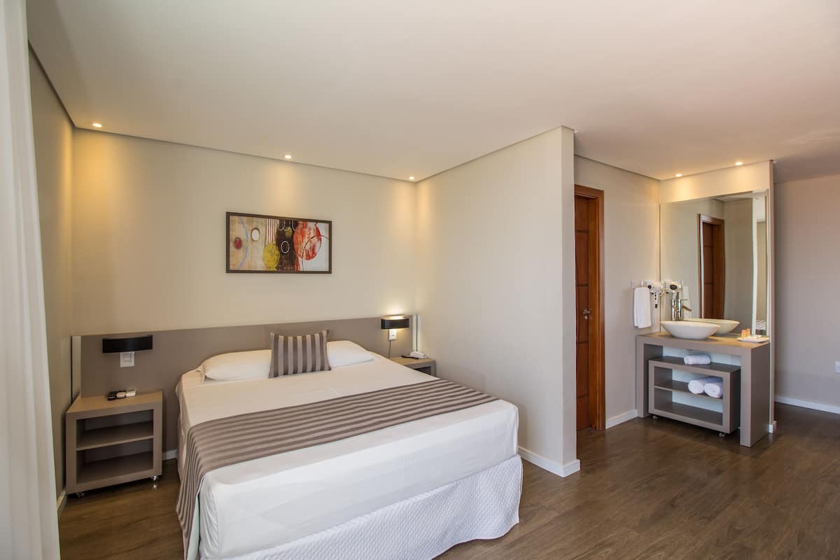 Quarto de hotel moderno com uma cama grande, mesas de cabeceira, banheiro com pia e espelho, e uma obra de arte pendurada na parede.