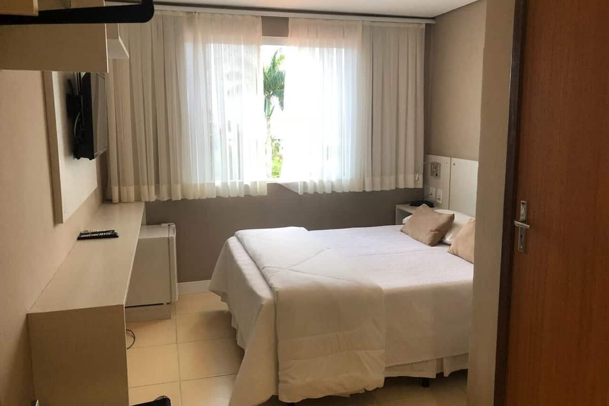 Quarto de hotel com cama, escrivaninha, cadeira, televisão e janela com cortinas.