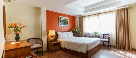 Quarto de hotel com uma cama, escrivaninha, cadeira, mesa de cabeceira, abajur e um quadro na parede.