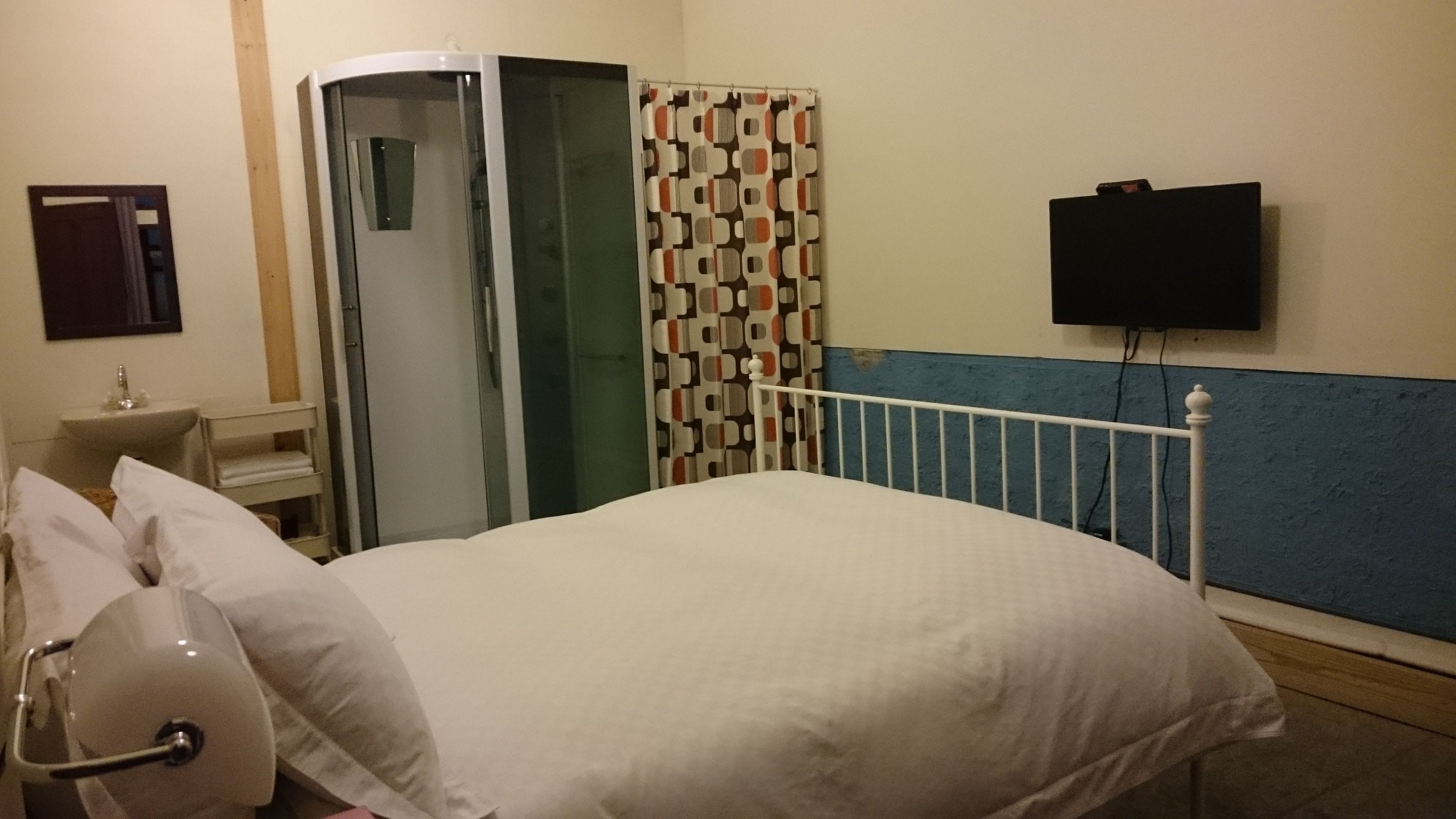 Quarto com cama, mesa de cabeceira, espelho, guarda-roupa, chuveiro e televisão.