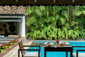Uma área à beira da piscina com espreguiçadeiras, uma mesa de jantar com pratos de comida e folhagem verde exuberante ao fundo.