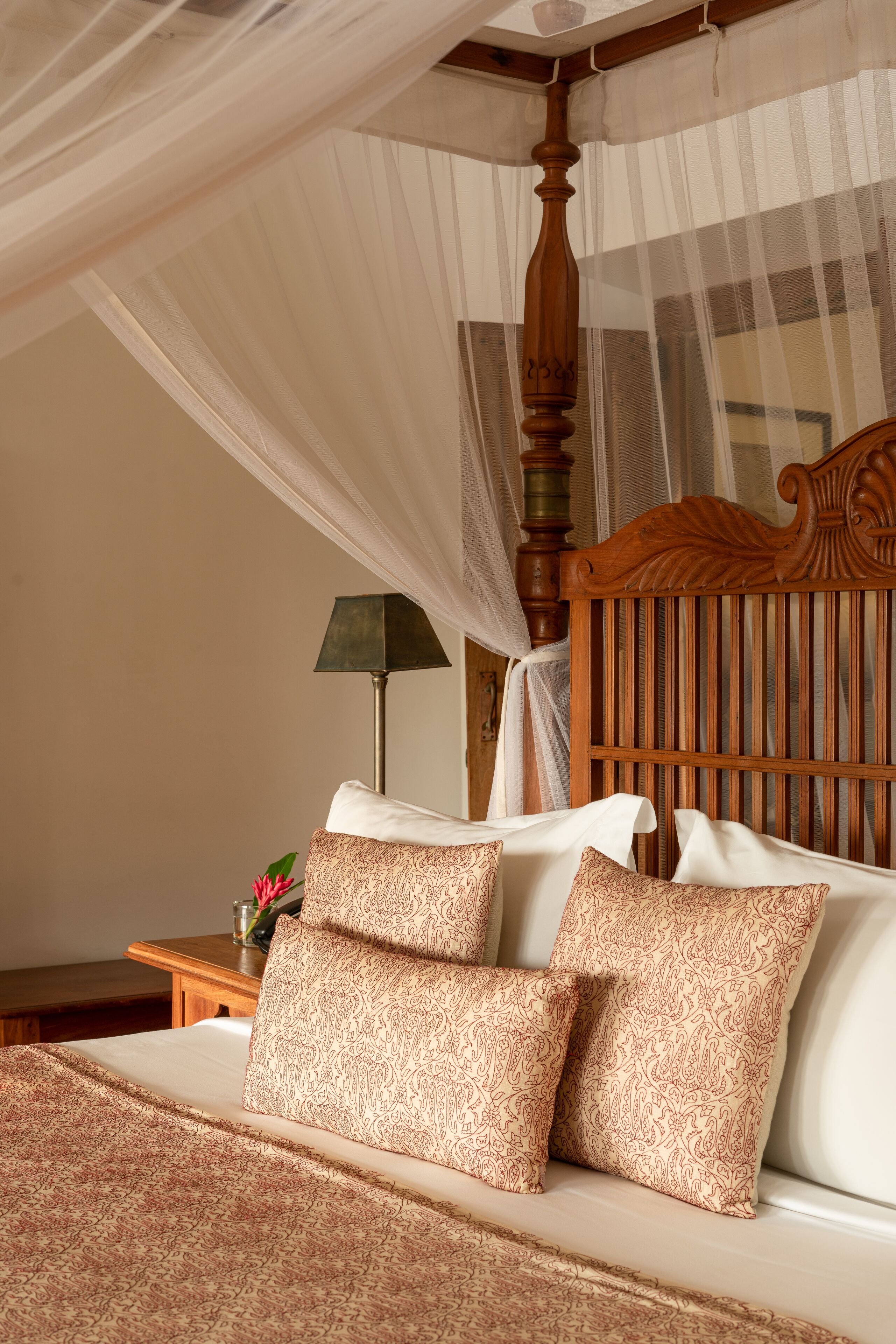 Quarto com cama de dossel, almofadas estampadas, cabeceira de madeira e mesa de cabeceira com abajur.
