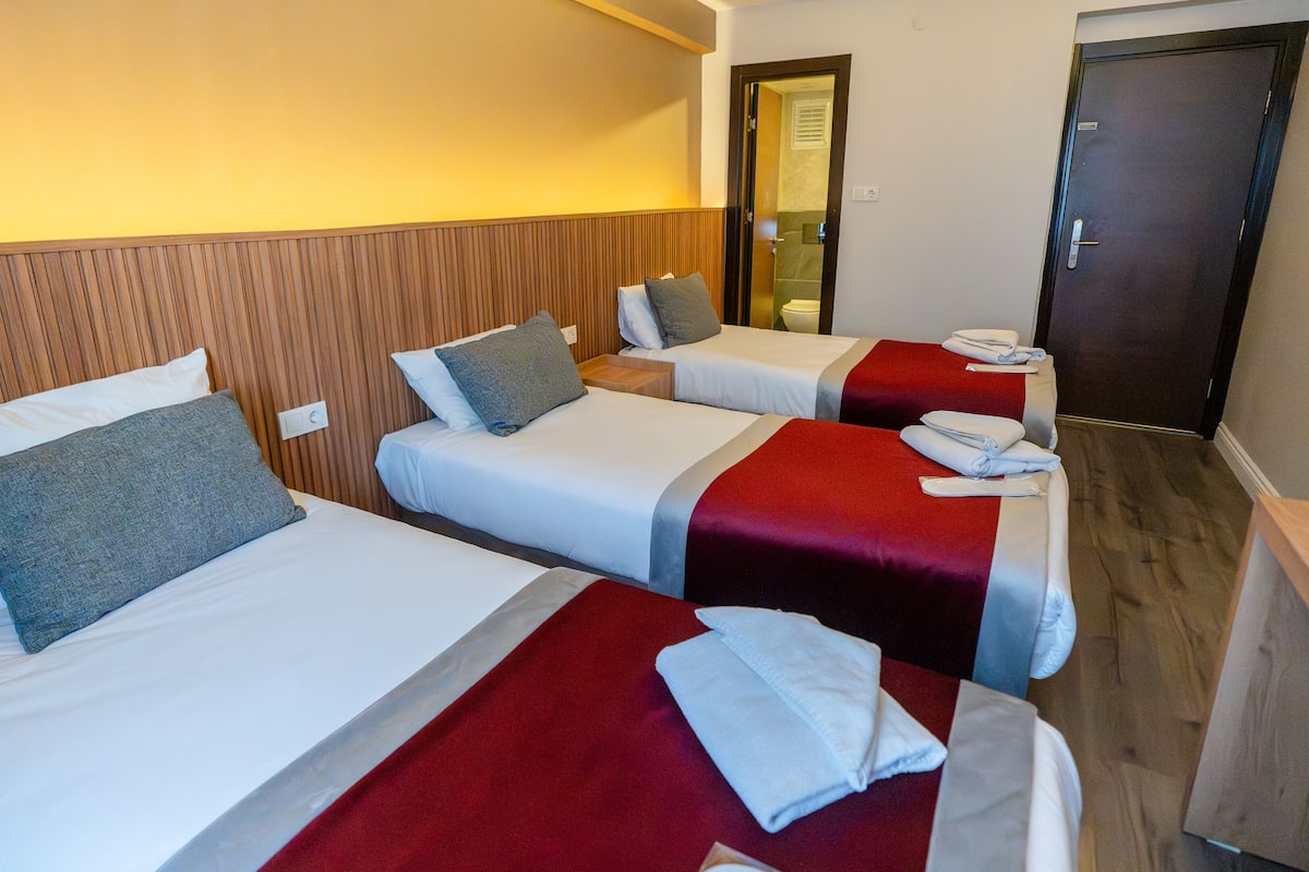 Quarto de hotel com três camas, cada uma com colcha vermelha e branca e almofadas cinzentas. Possui cabeceira, mesa de cabeceira e escrivaninha de madeira, esta última com uma cadeira. O chão é de madeira e há uma porta à direita.