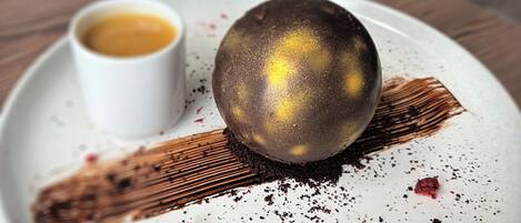 Sobremesa em um prato branco, com uma esfera de chocolate, um traço de pincel de chocolate e raspas de chocolate espalhadas.