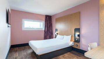 Quarto de hotel com cama, escrivaninha, janela com cortinas e televisão presa à parede.