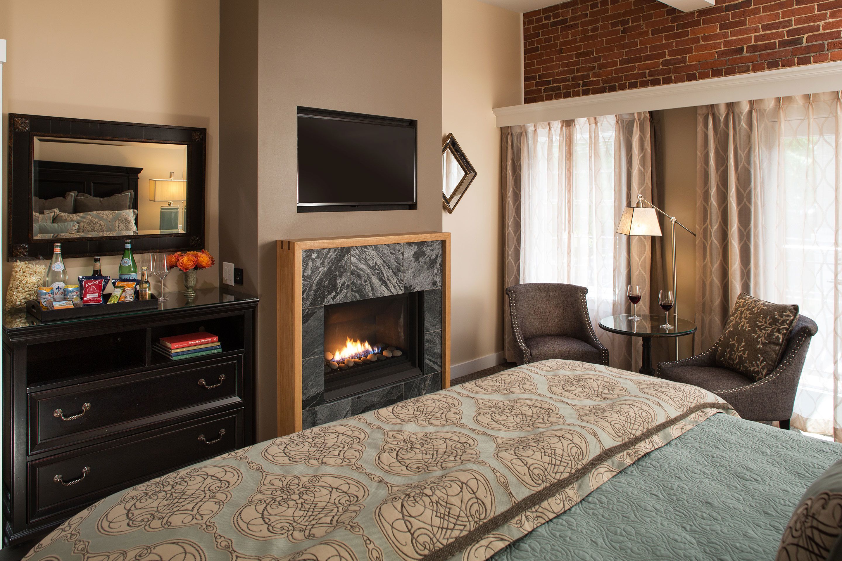 Quarto de hotel com lareira, cama com roupa de cama estampada, televisão e área de estar com abajur e poltrona.