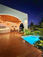 Uma área de estar moderna ao ar livre, com um deck de madeira, piscina e um espaço de convívio com parede branca e teto de madeira.