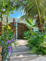 Uma trilha de madeira serpenteia por um jardim exuberante com plantas tropicais e um muro de pedra.