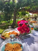 Uma área de refeições ao ar livre com uma mesa posta para o café da manhã, incluindo waffles, frutas e um prato tampado.