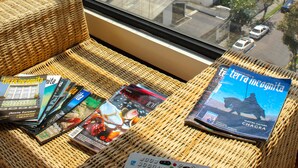 Um sofá de vime com revistas e um controle remoto, com vista para o horizonte urbano através de uma janela.