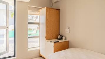 Quarto com cama, guarda-roupa de madeira, pequena mesa com chaleira e chávena, e janela com vista para o exterior.