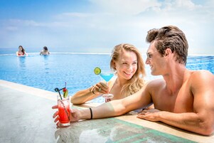 Um casal relaxando à beira da piscina com bebidas, com vista para o oceano ao fundo.