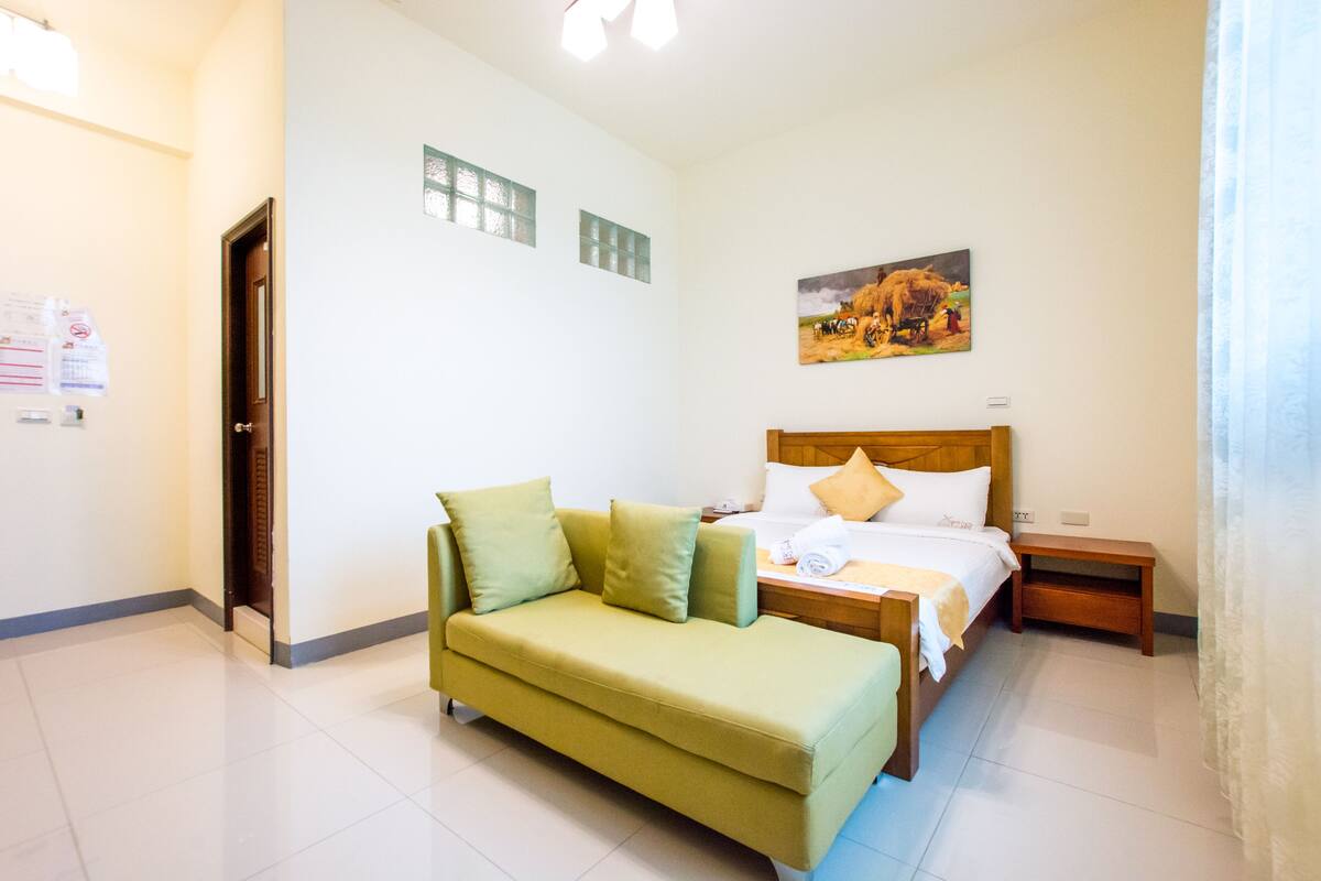 Quarto de hotel com cama, sofá verde, mesa de cabeceira de madeira e um quadro na parede.